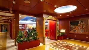 Um hall de hotel com piso de mármore, decoração em vermelho e dourado, e uma pintura grande na parede.