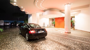 Um carro preto estacionado em um caminho de paralelepípedos à noite, com a entrada de um prédio moderno que apresenta um telhado curvo e luminárias no teto.