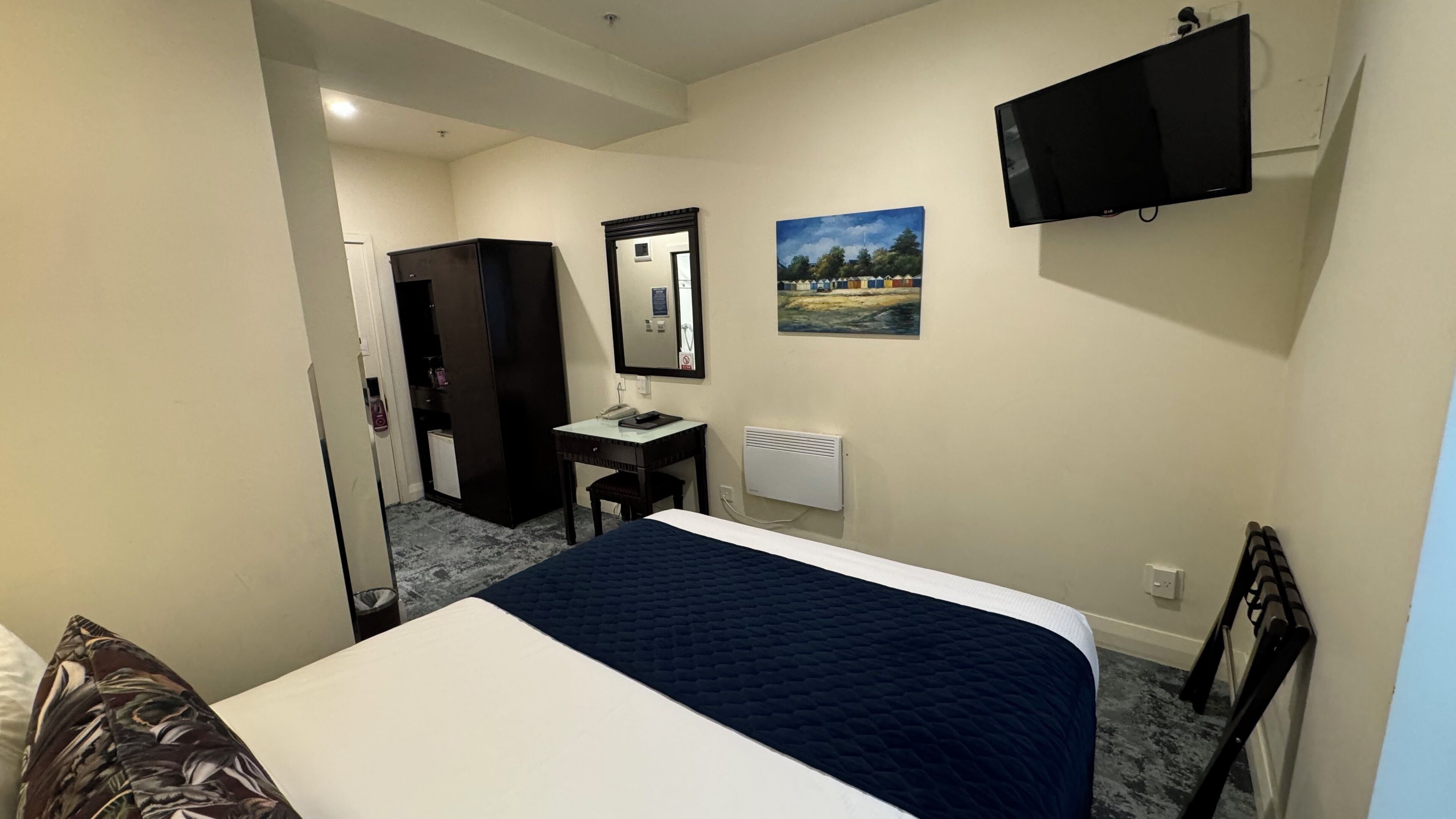 Quarto de hotel com cama, escrivaninha, cadeira, espelho, quadro, televisão e radiador.