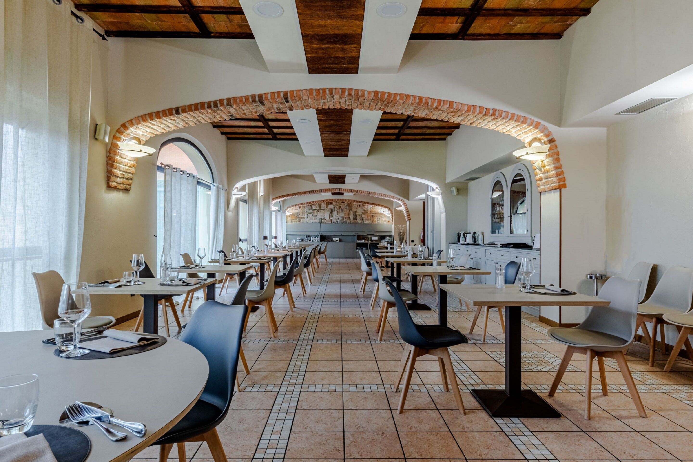 Um restaurante com várias mesas e cadeiras, uma cozinha ao fundo e piso ladrilhado.