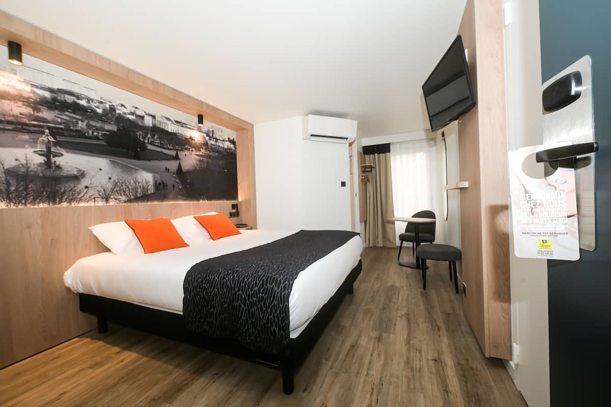 Quarto de hotel com cama, escrivaninha, cadeira, TV fixada na parede e um cartaz.