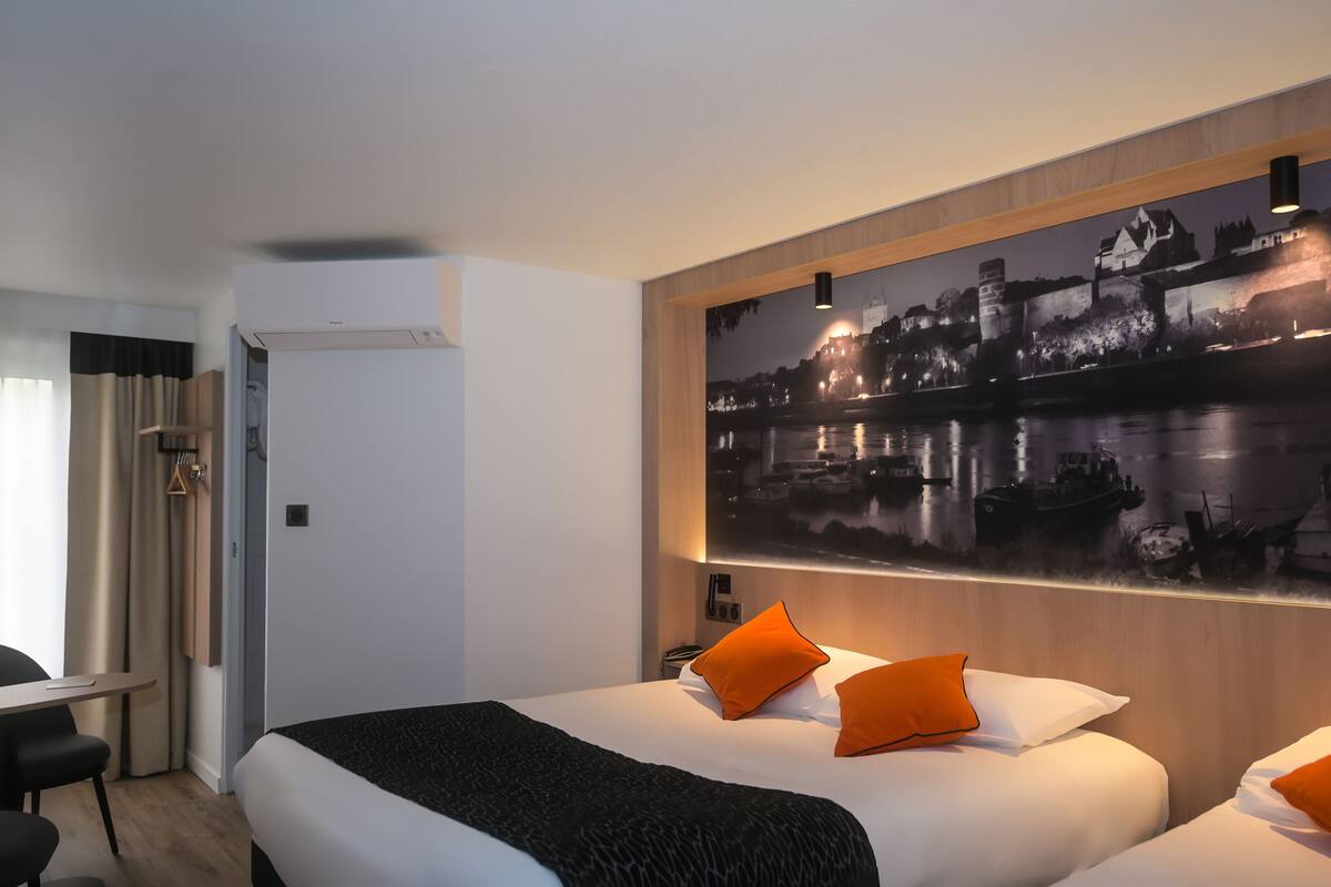 Quarto de hotel com uma cama grande, duas almofadas laranja, um cobertor preto e um quadro pendurado na parede com uma paisagem urbana noturna.