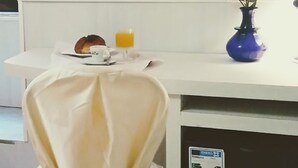 Uma cadeira com capa bege amarrada atrás, uma pequena mesa com um croissant, uma xícara e um copo de suco de laranja, e um vaso com flores em um balcão.