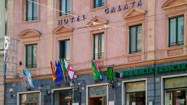 Um hotel chamado 'HOTEL GALATA' com várias bandeiras, incluindo a bandeira da União Europeia, e uma farmácia chamada 'FARMACIA FRASCAR' do outro lado da rua.
