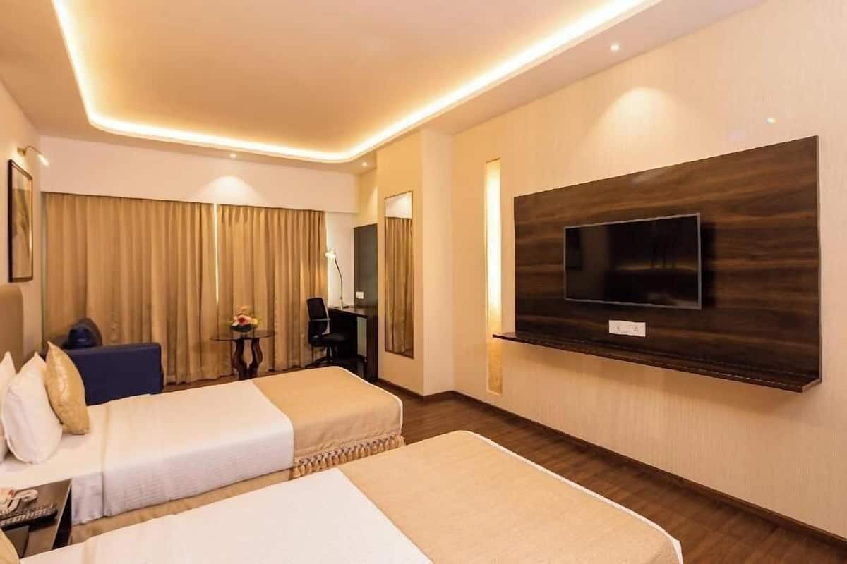 Quarto de hotel com duas camas, uma escrivaninha, uma cadeira, televisão na parede e uma janela grande com cortinas.