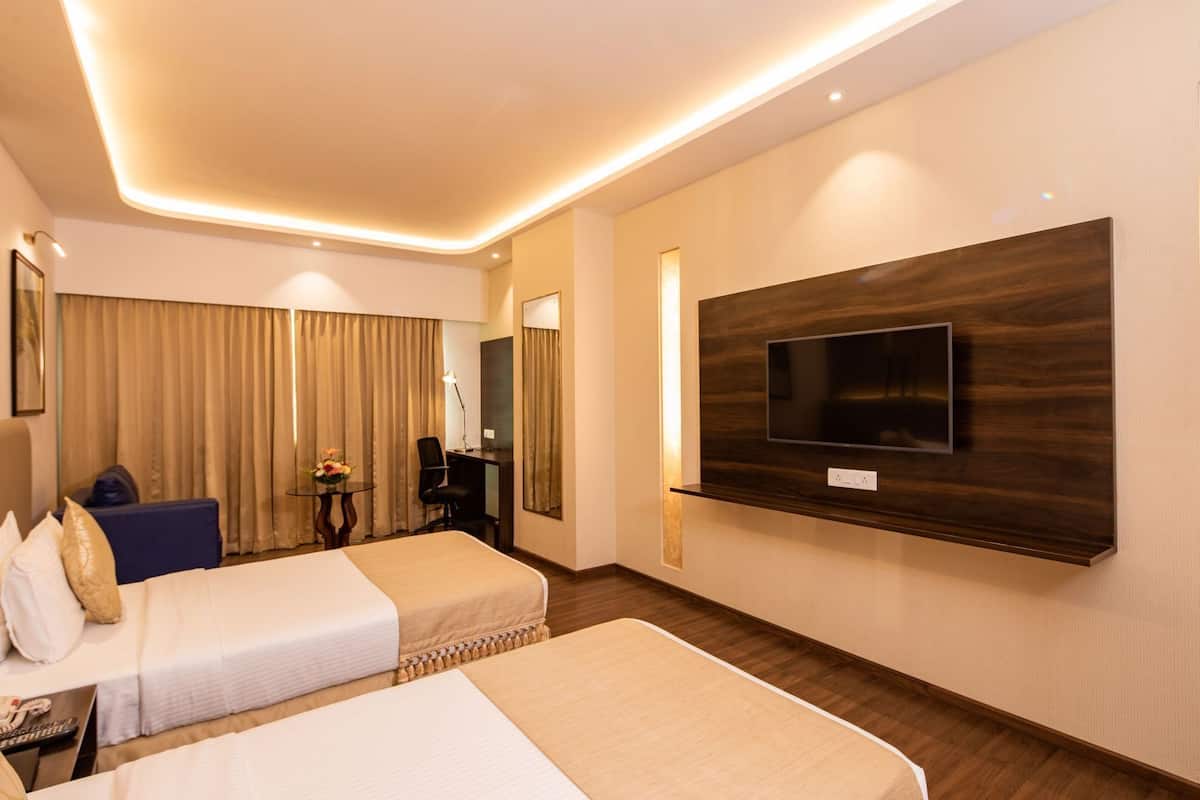 Quarto de hotel com duas camas, uma escrivaninha, uma cadeira, televisão na parede e uma janela grande com cortinas.