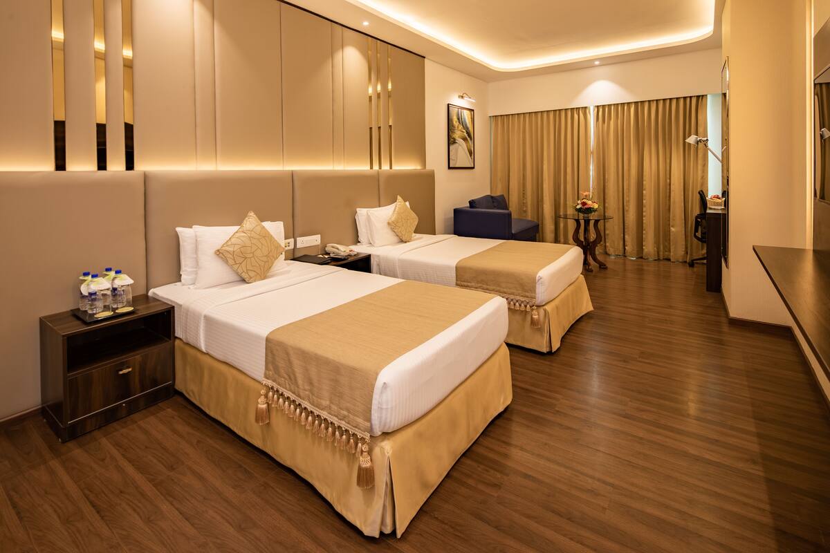 Quarto de hotel com duas camas, uma mesa, uma cadeira, uma mesinha de cabeceira e uma pequena mesa com abajur.