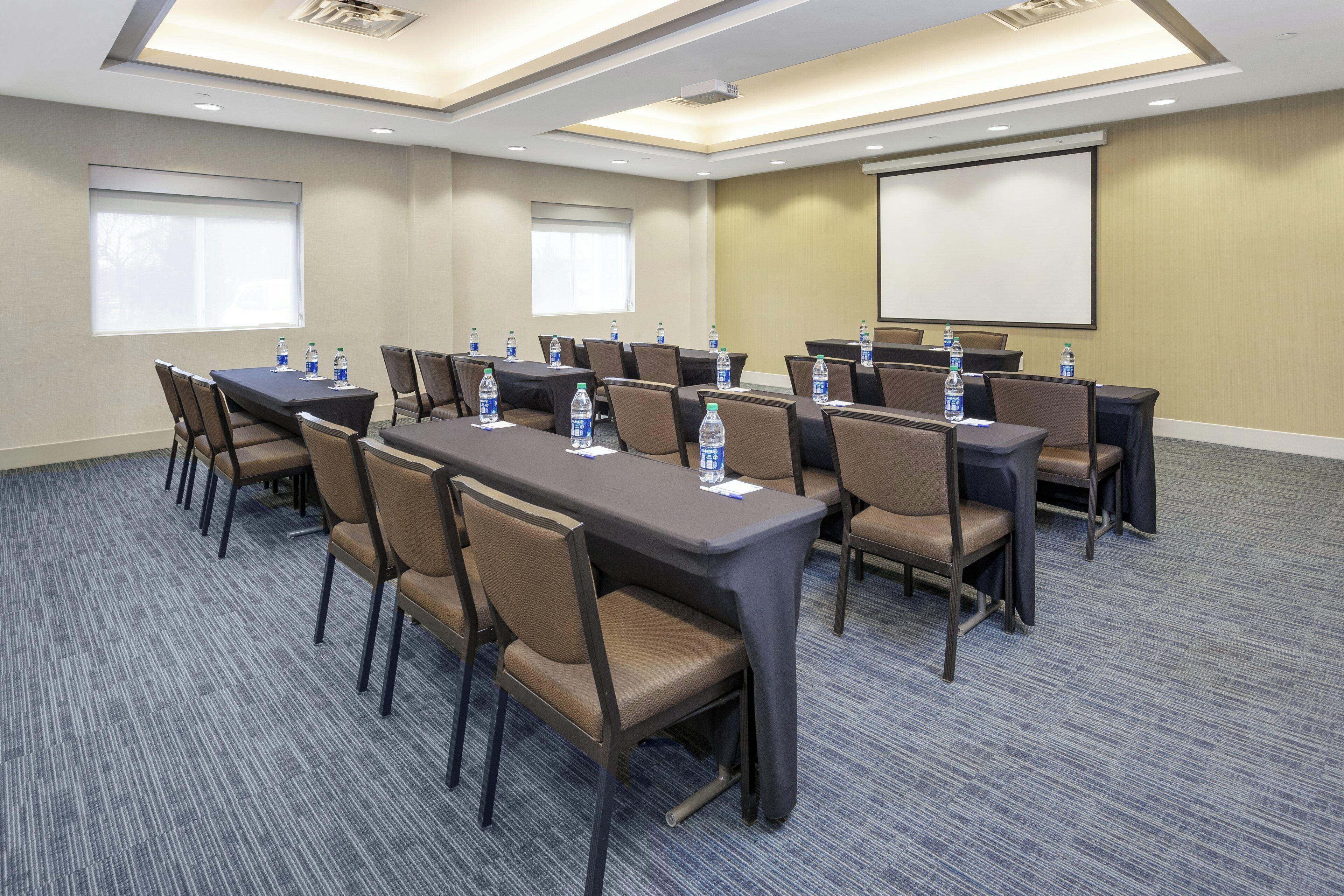 Sala de conferências com várias mesas e cadeiras, equipada com garrafas de água, tela de projetor e uma janela ampla.