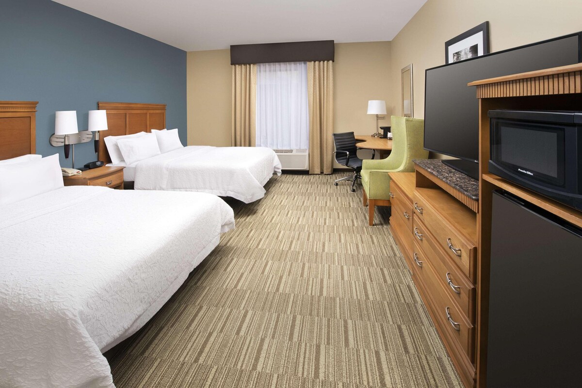 Quarto de hotel com duas camas, uma escrivaninha com cadeira, televisão e minibar.