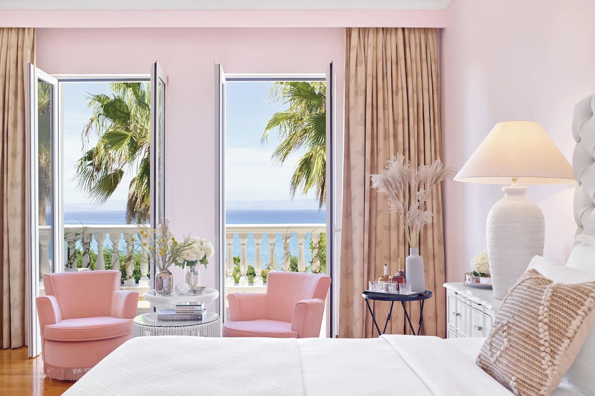 Quarto com uma cama grande, duas cadeiras cor-de-rosa, uma mesa redonda com um vaso e vista para o mar através de cortinas abertas.