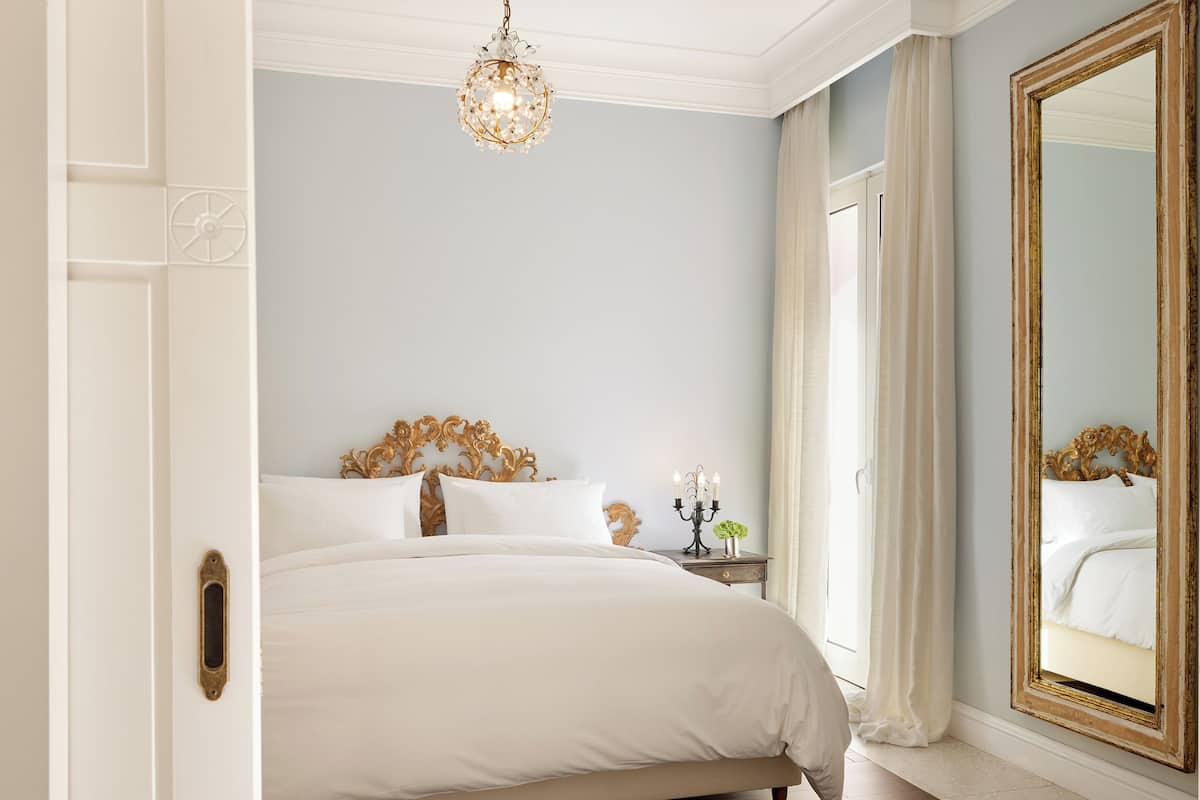 Quarto com cama grande, lustre, mesa de cabeceira com castiçal e espelho com moldura dourada.