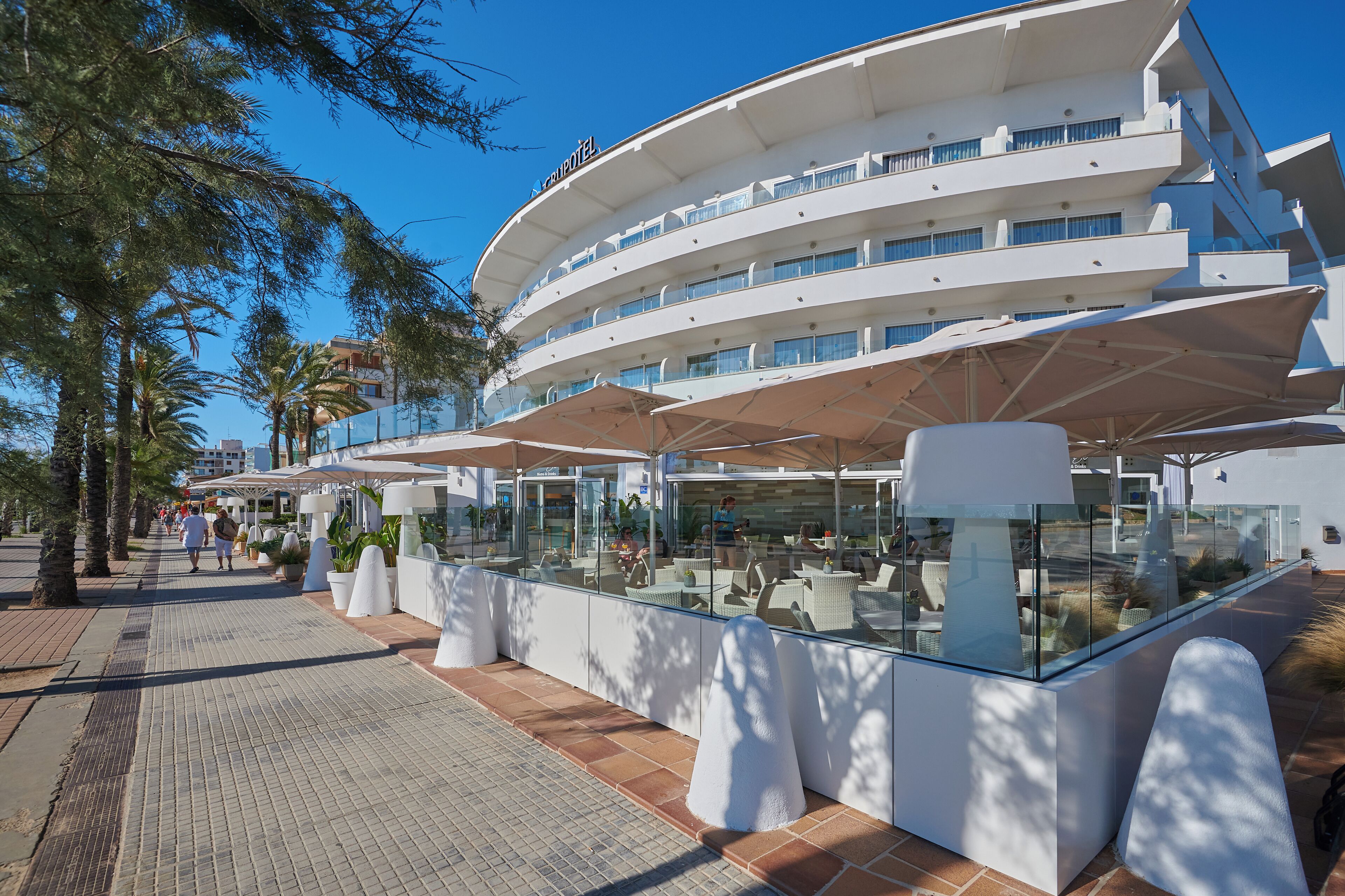 Um hotel de vários andares com áreas de estar ao ar livre, um terraço coberto e vista clara das palmeiras ao redor.