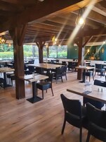 Restaurante com mesas e cadeiras de madeira, janelas amplas e luminárias pendentes.