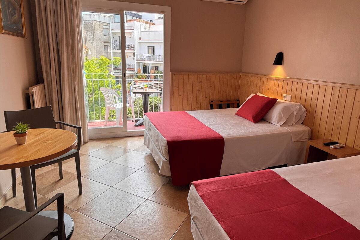 Quarto de hotel com cama, uma pequena mesa com cadeira, planta em vaso e varanda com vista para edifícios e vegetação.