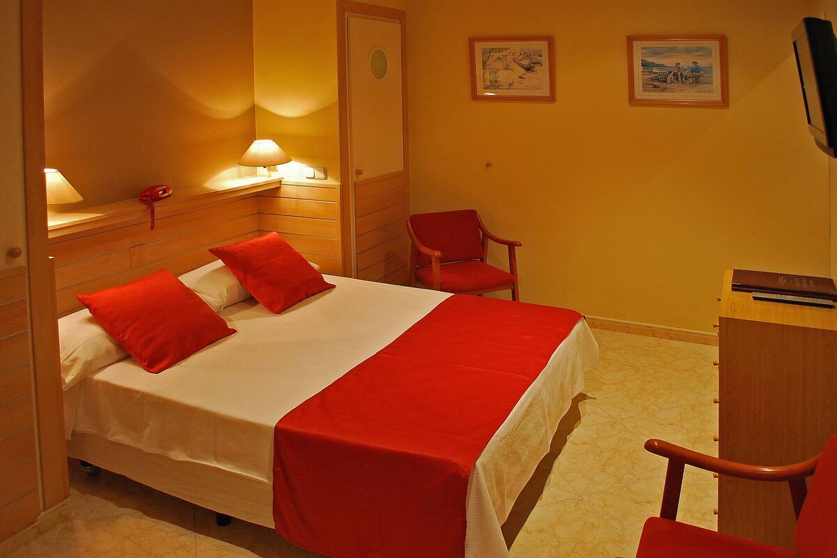 Quarto de hotel com cama, uma cadeira, escrivaninha e televisão na parede.