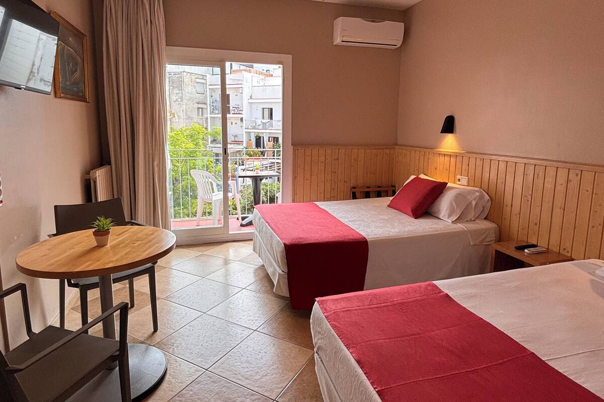 Quarto de hotel com duas camas, uma mesinha com uma cadeira, televisão na parede e varanda com vista para edifícios.