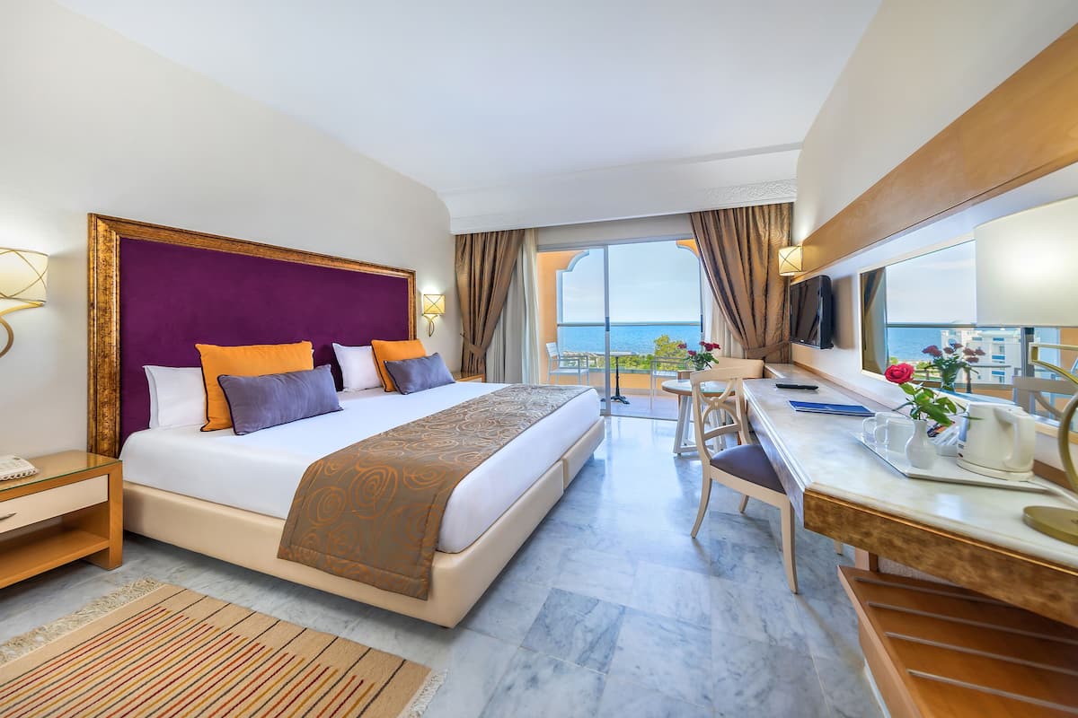 Quarto de hotel com uma cama grande, uma escrivaninha com cadeira, vista para o mar e uma varanda com mesa e cadeiras.
