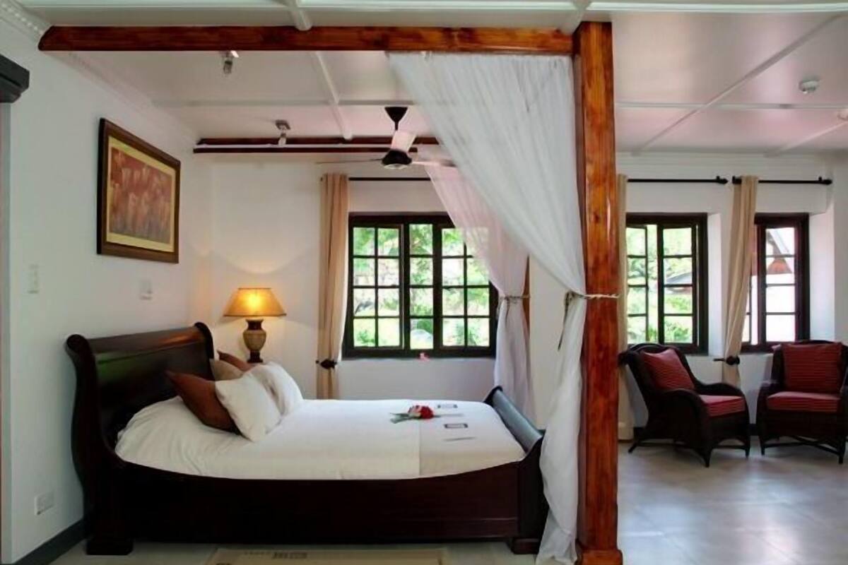 Quarto com cama, mesa de cabeceira, luminária e janela com cortinas.