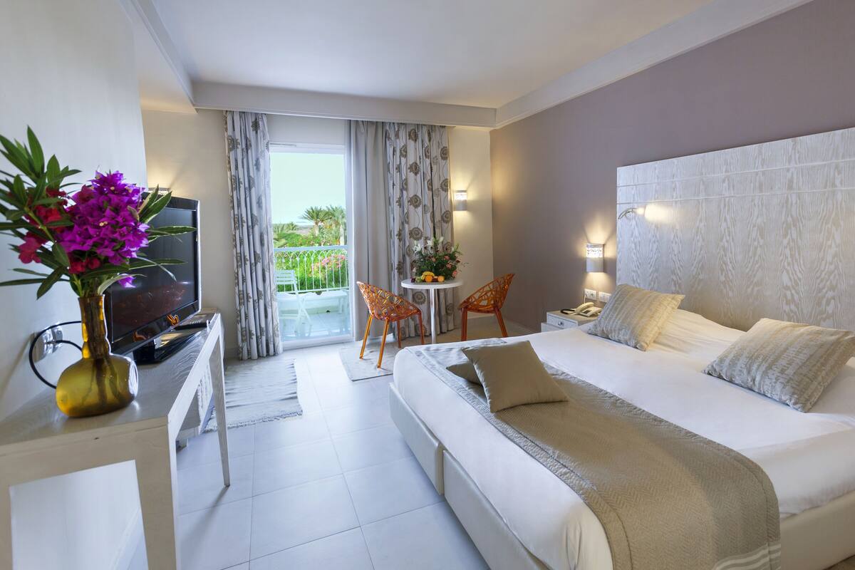 Quarto de hotel com cama, escrivaninha com televisão, varanda com mesa e cadeiras, e um vaso com flores.