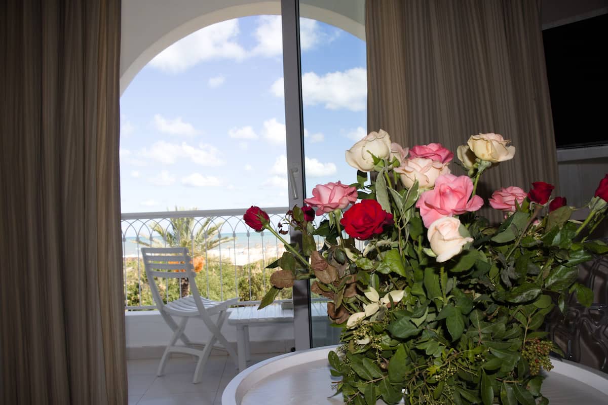 Uma varanda com uma cadeira branca, uma mesa com um vaso de rosas e vista para a praia e palmeiras.