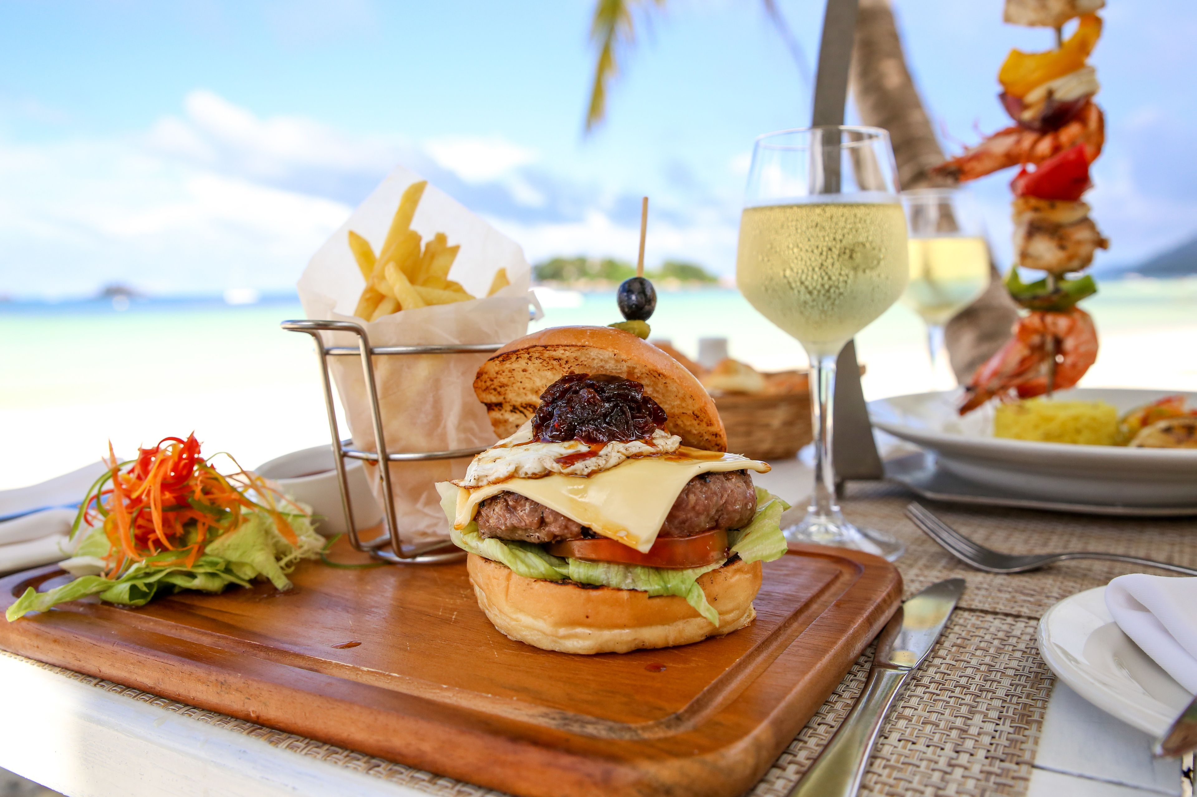 Uma refeição com hambúrguer, batatas fritas e uma bebida sobre uma tábua de madeira, com vista para o oceano ao fundo.