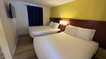 Quarto de hotel com duas camas, televisão fixada na parede, mesa de cabeceira com abajur e cabeceira de madeira.