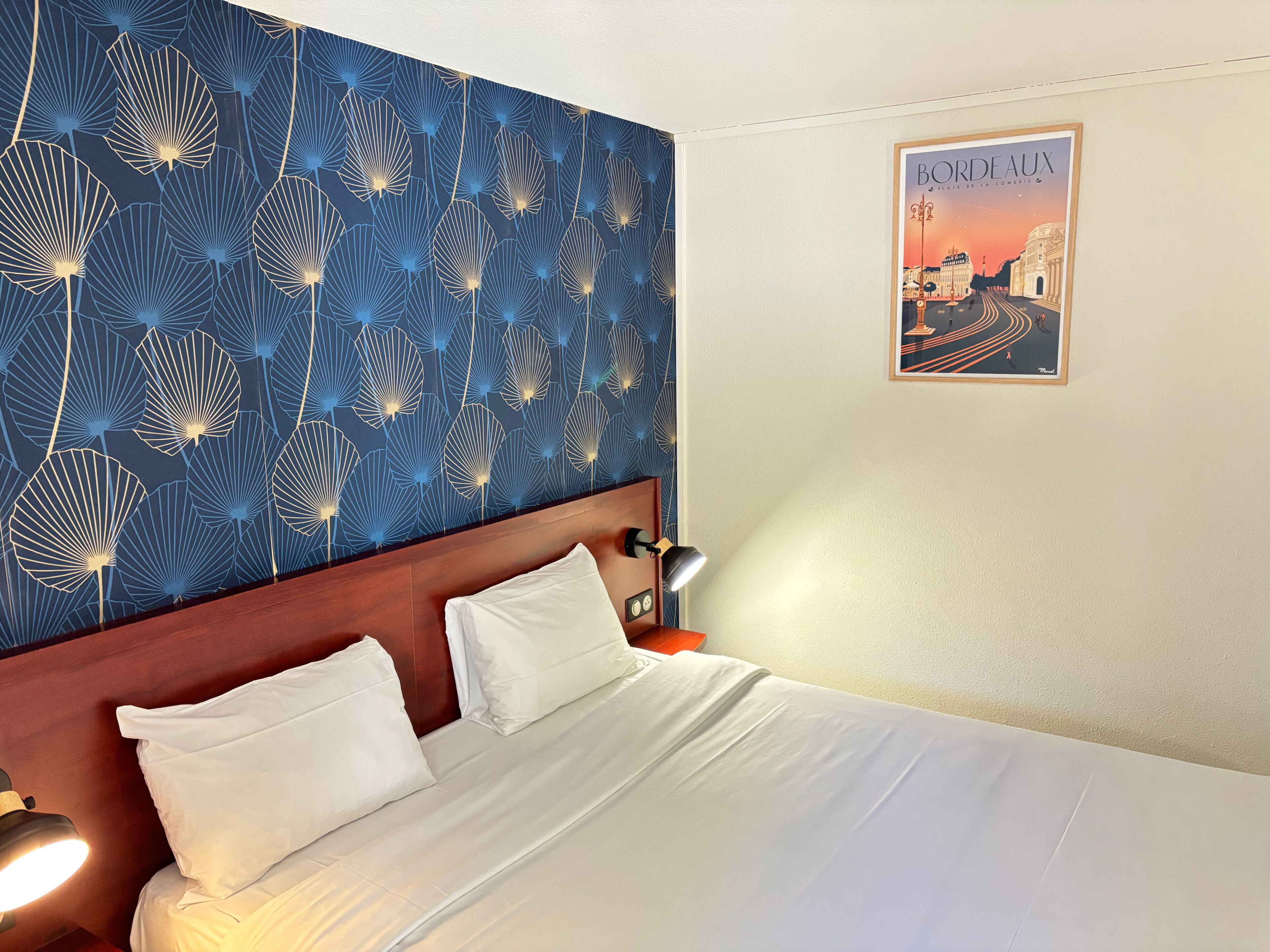 Quarto de hotel com cama, almofadas brancas, cabeceira, parede com papel de parede floral azul, mesa de cabeceira, abajur e um cartaz de Bordeaux.