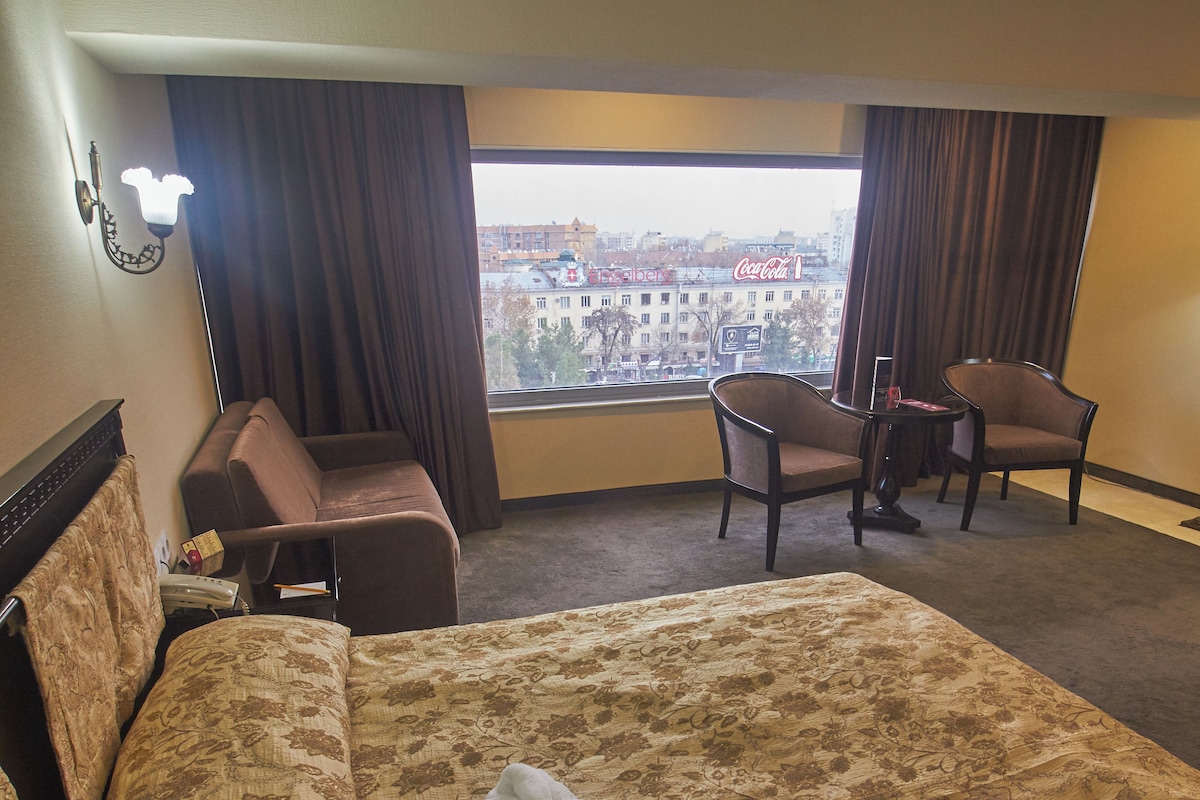 Quarto de hotel com cama, sofá, duas cadeiras, uma mesa pequena e vista para a cidade através de amplas janelas.