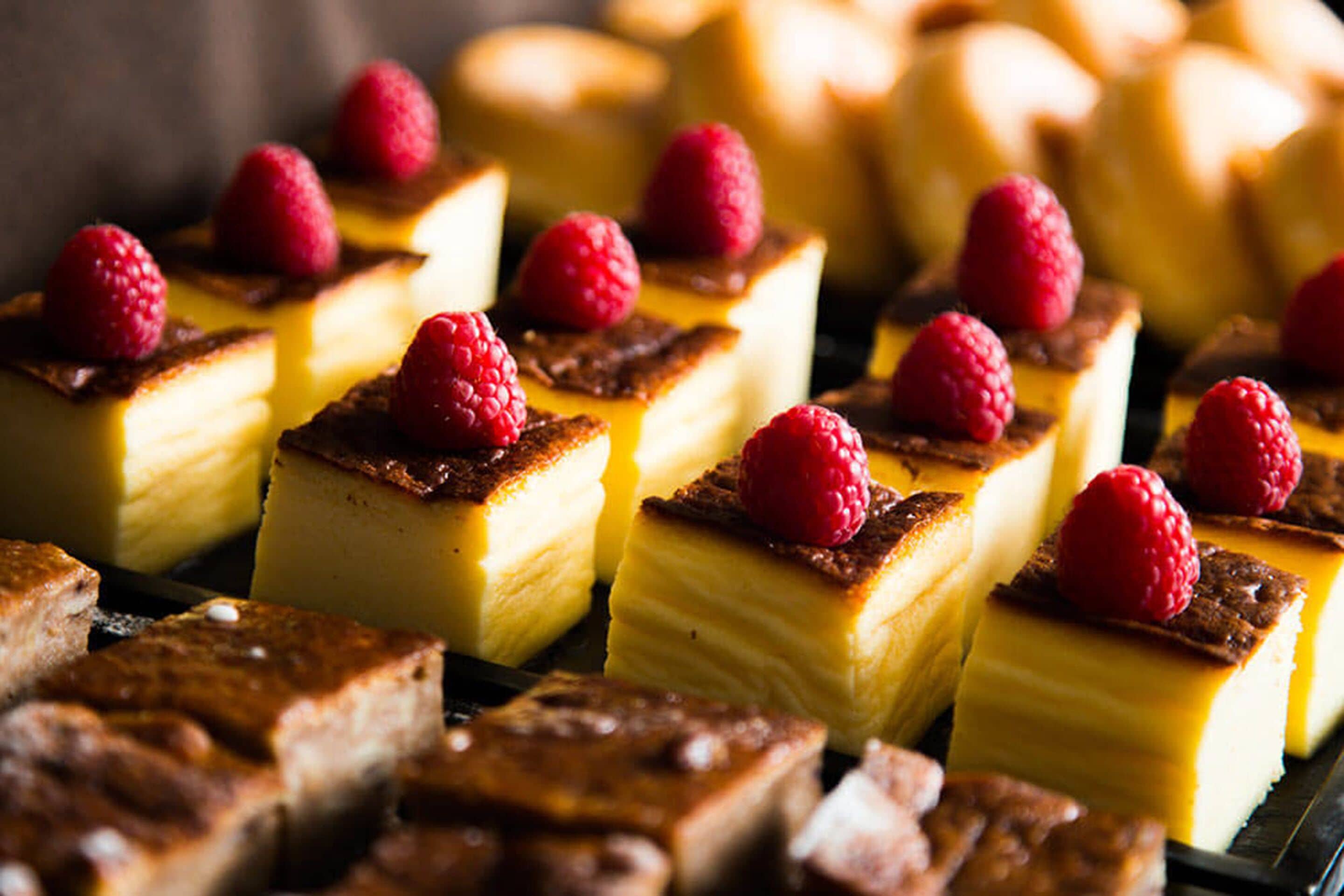Diversos doces quadrados com cobertura dourada, alguns adornados com framboesas.