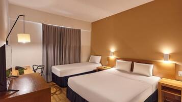 Quarto de hotel com duas camas, uma escrivaninha com luminária, uma cadeira e uma janela com cortinas.