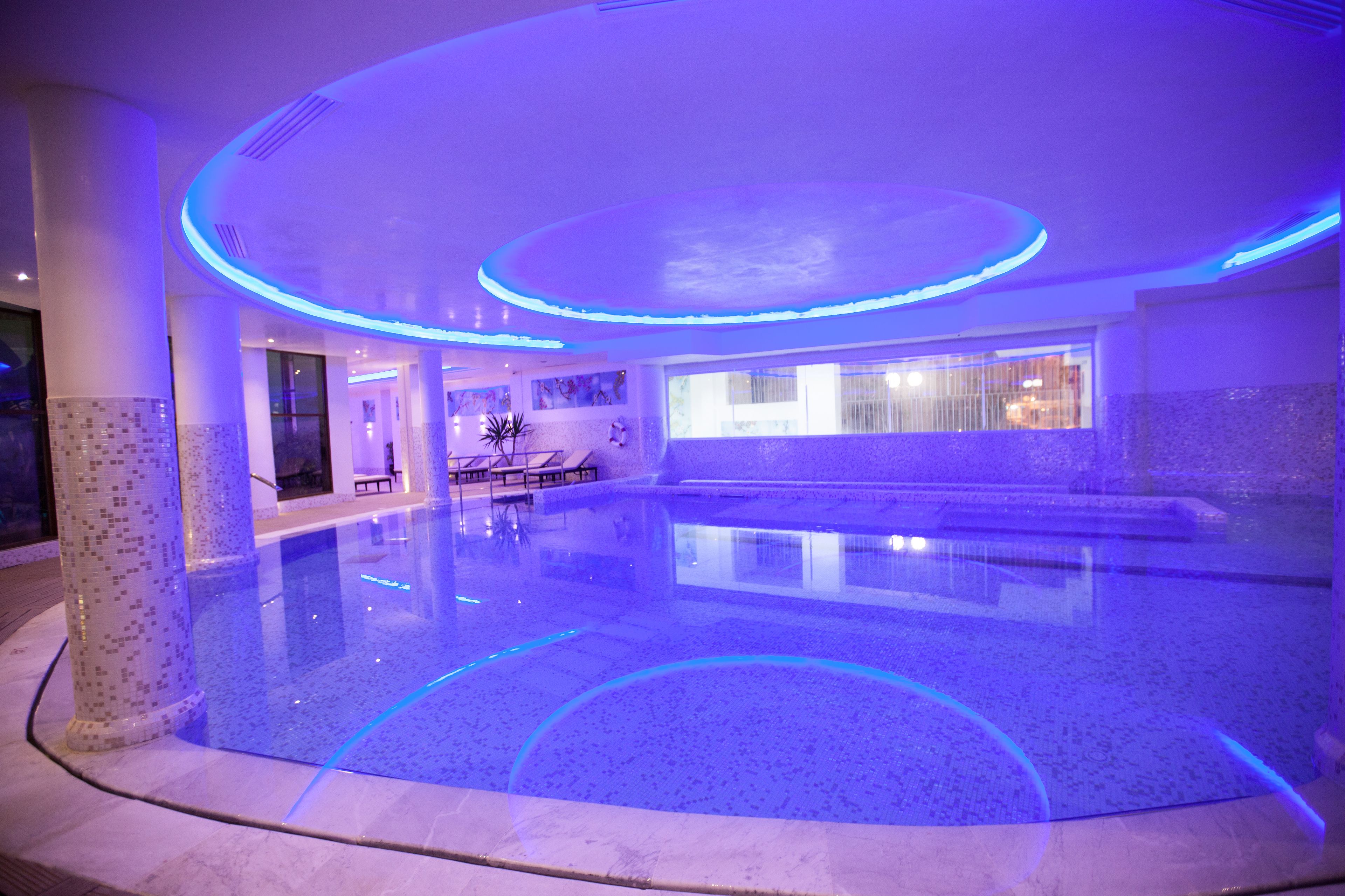 Uma grande piscina interna com iluminação azul, área de estar e vista para o exterior.