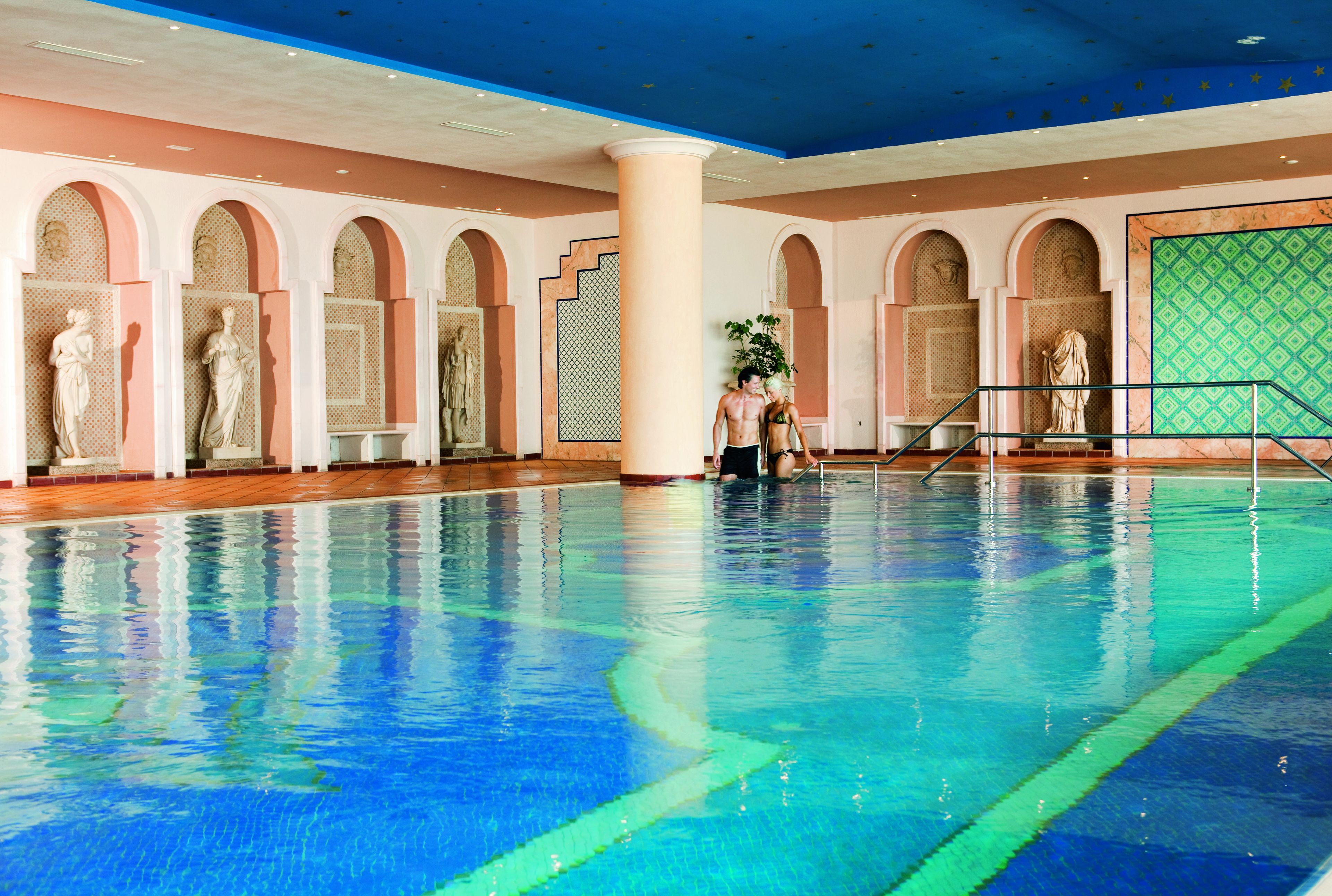 Uma grande piscina interna com água azul cristalina, ladeada por colunas em arco e estátuas.