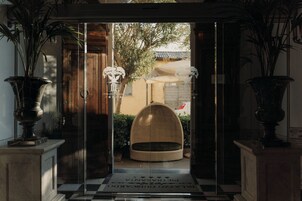 Entrada de hotel com chão xadrez, uma cadeira de vime e plantas em vasos.