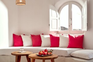 Quarto com sofá branco, almofadas vermelhas e brancas, mesa de madeira com um copo de água e uma bandeja de frutas, e janela com design em arco.
