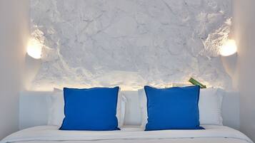 Cama com roupa de cama branca e duas almofadas azuis, parede branca texturizada ao fundo, e duas luzes fixadas na parede.