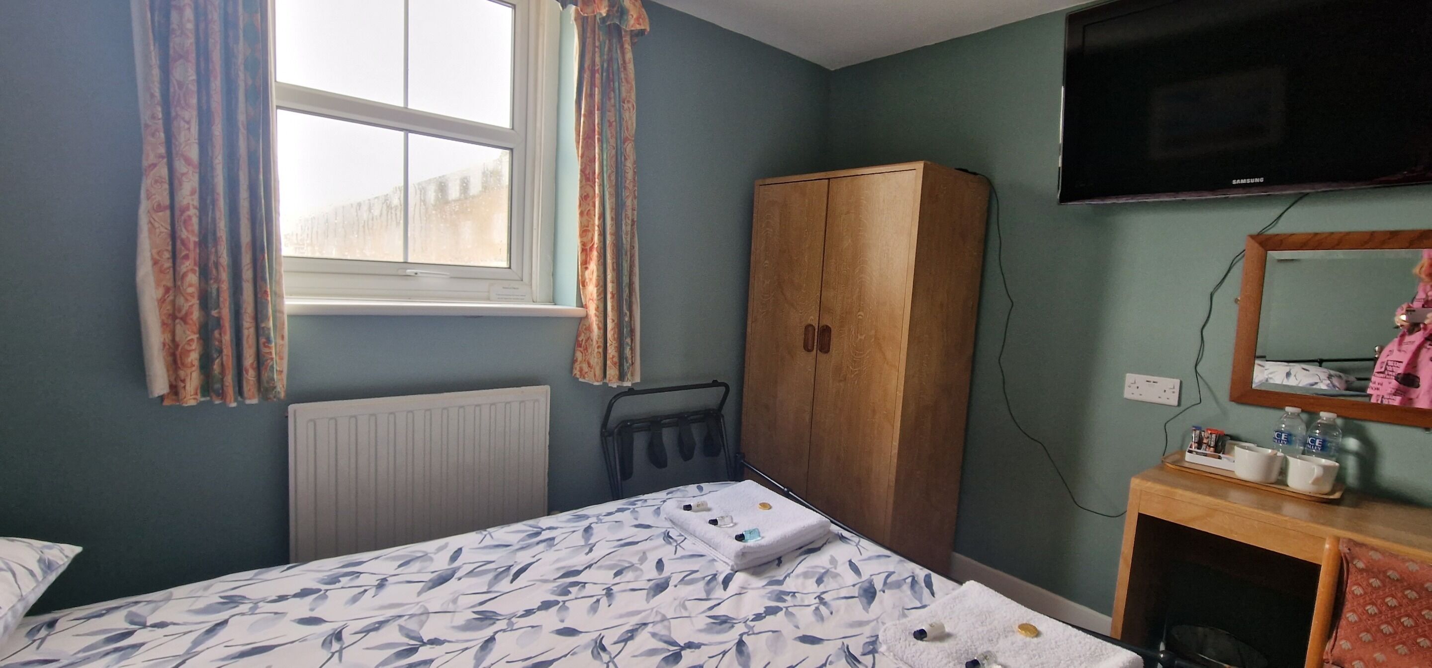Quarto com cama, guarda-roupa, radiador, televisão e janela com cortinas.