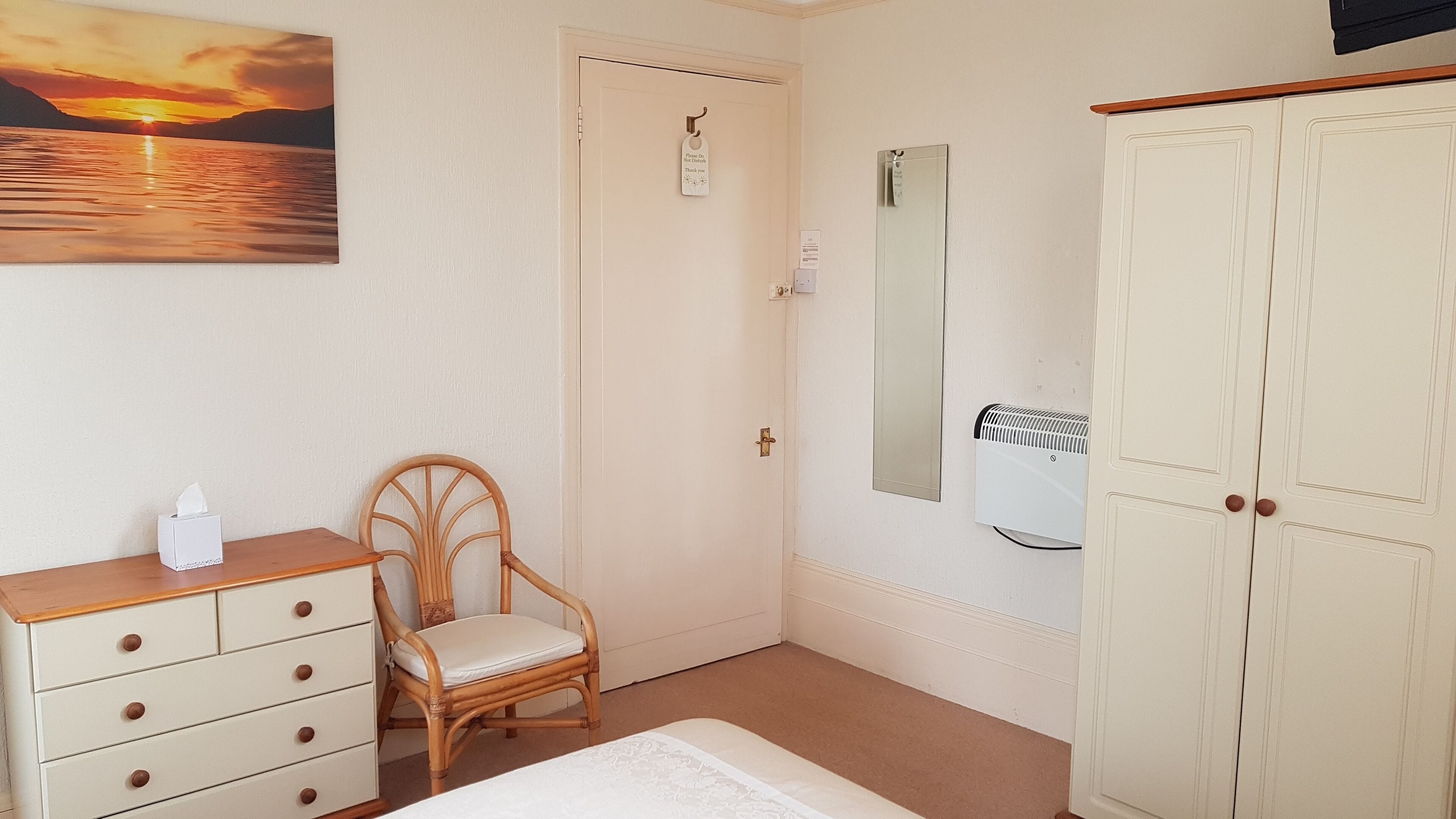 Um quarto com cama, cómoda de madeira, cadeira de vime, guarda-roupa, aquecedor fixado na parede e um quadro de um pôr do sol sobre a água.