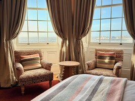 Quarto com duas poltronas estampadas, uma mesa redonda pequena e uma cama com manta listrada.