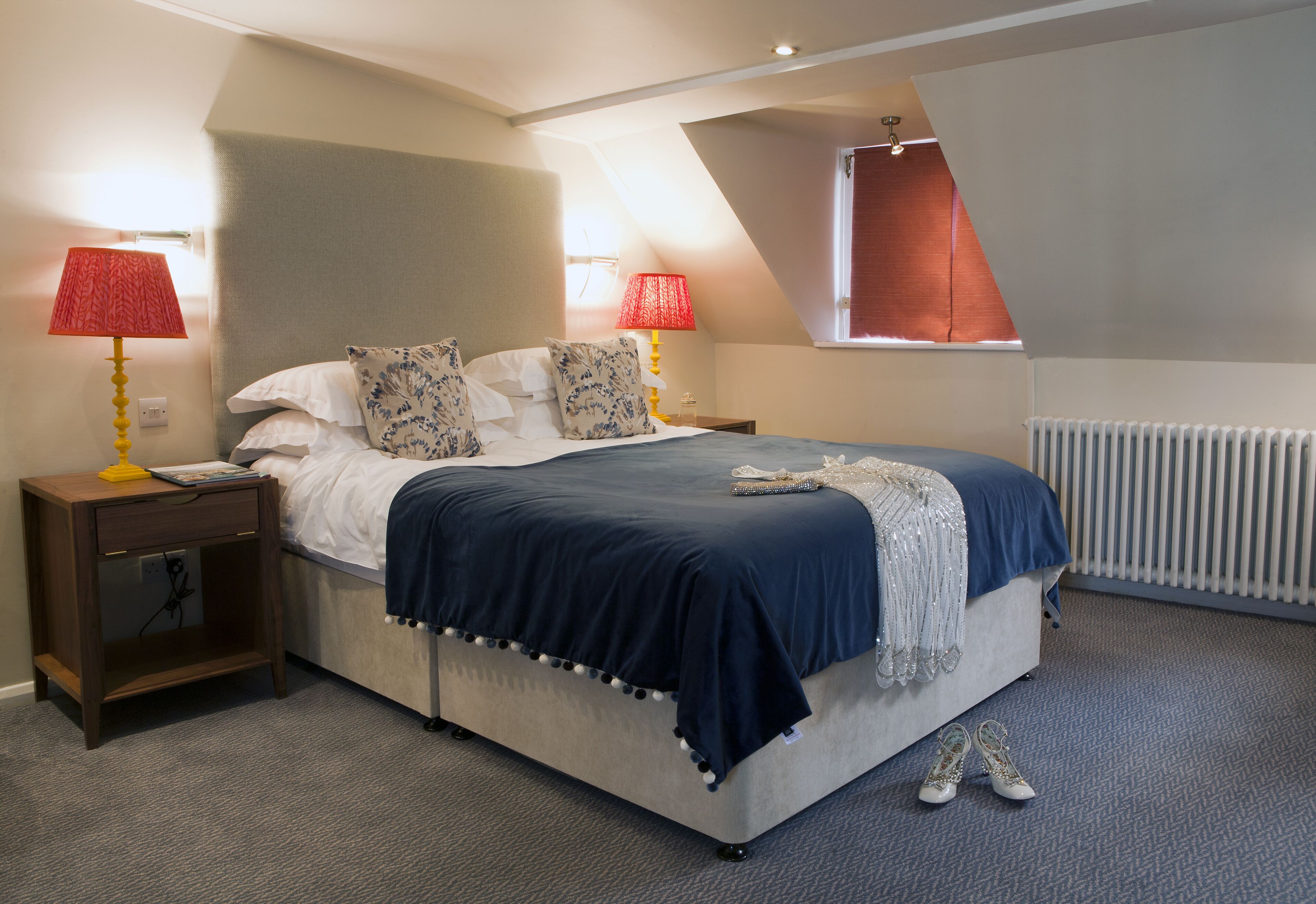 Quarto com cama, duas mesas de cabeceira, luminárias, radiador e um par de sapatos no chão.