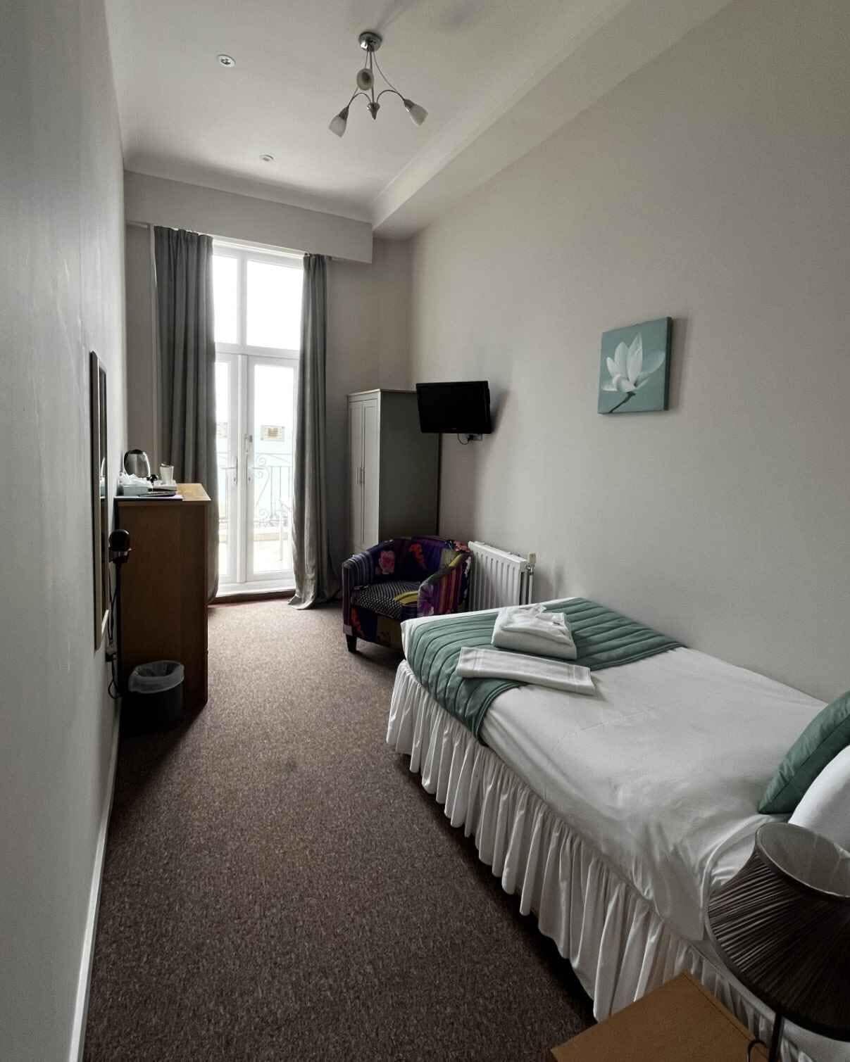 Quarto de hotel com cama, escrivaninha com cadeira, radiador, televisão na parede e janela com cortinas.