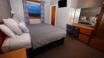 Quarto com cama, escrivaninha, cadeira, espelho, televisão e janela com cortinas.