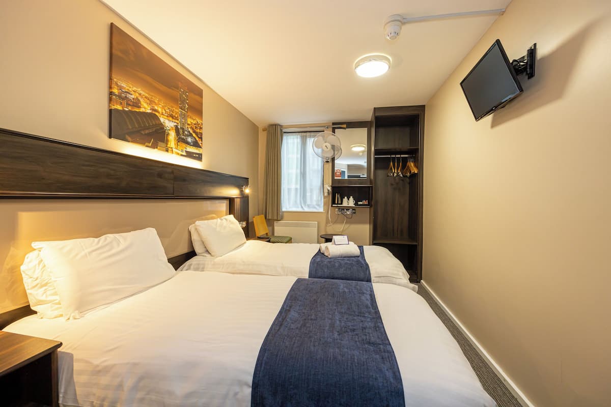 Quarto de hotel com duas camas, uma televisão de tela plana na parede, uma janela com cortinas e um cabideiro com roupas penduradas.
