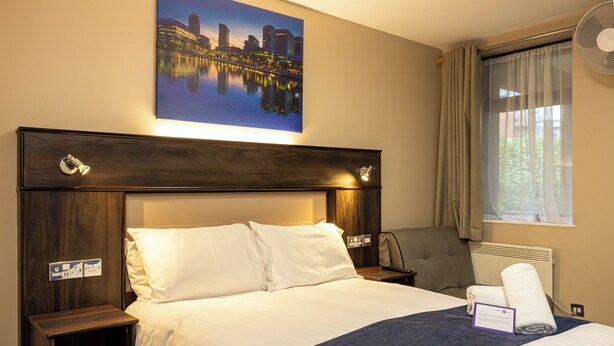 Quarto de hotel com uma cama grande, mesa de cabeceira, cadeira, janela com cortinas e uma pintura da paisagem urbana na parede.