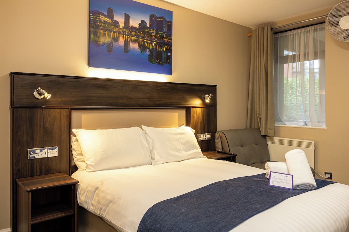 Quarto de hotel com uma cama grande, mesa de cabeceira, cadeira, janela com cortinas e uma pintura da paisagem urbana na parede.