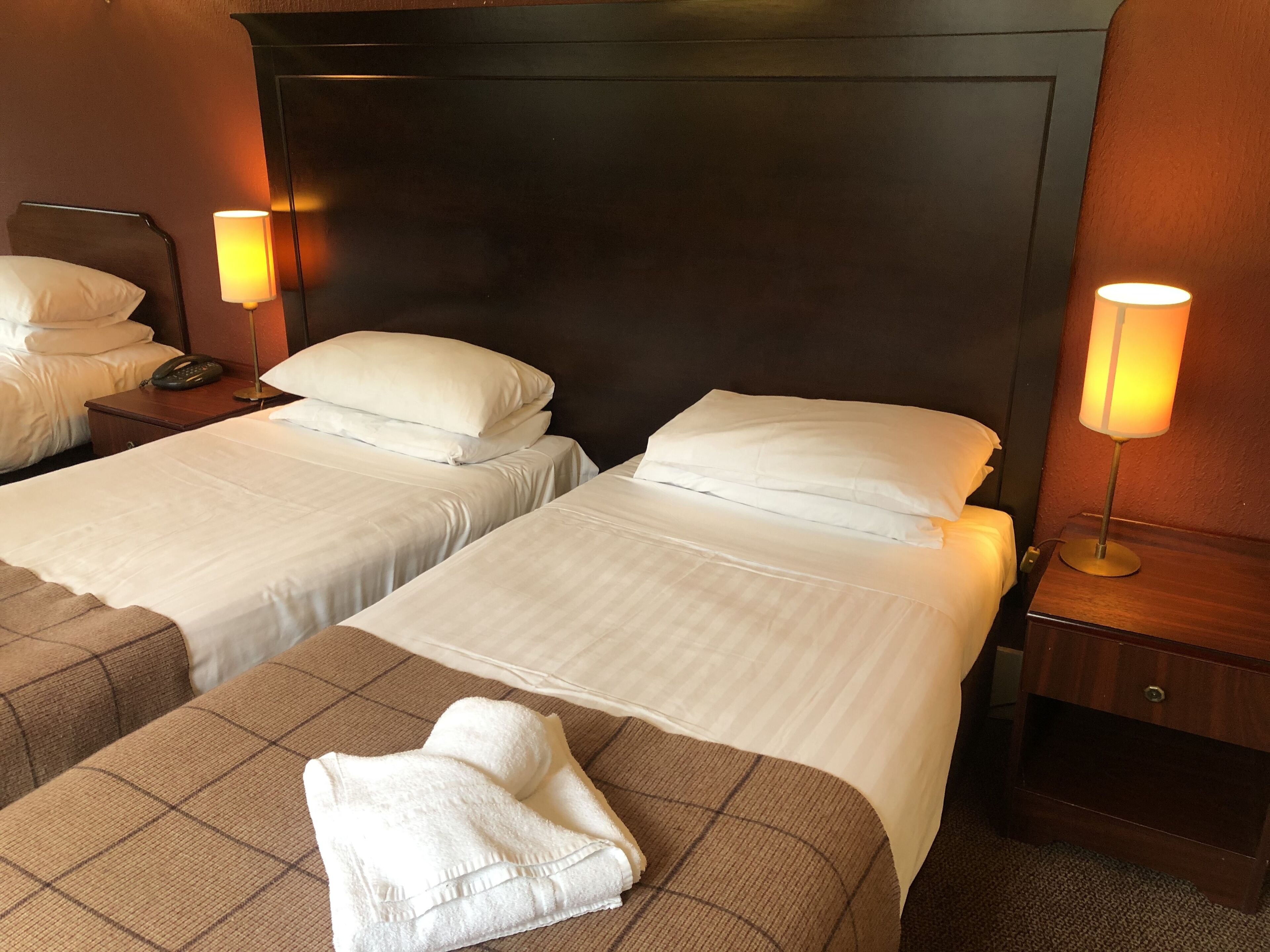 Quarto de hotel com duas camas, cada uma com roupa de cama e almofadas brancas, cabeceira de madeira, luminárias laterais, telefone e uma pequena mesa de cabeceira de madeira com gaveta.