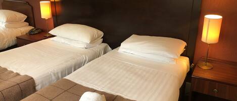 Quarto de hotel com duas camas, cada uma com roupa de cama e almofadas brancas, cabeceira de madeira, luminárias laterais, telefone e uma pequena mesa de cabeceira de madeira com gaveta.