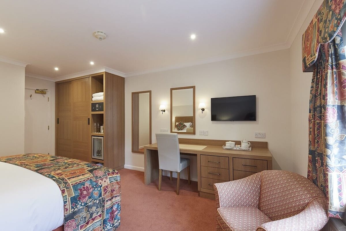Quarto de hotel com cama, escrivaninha, cadeira, televisão e guarda-roupa.