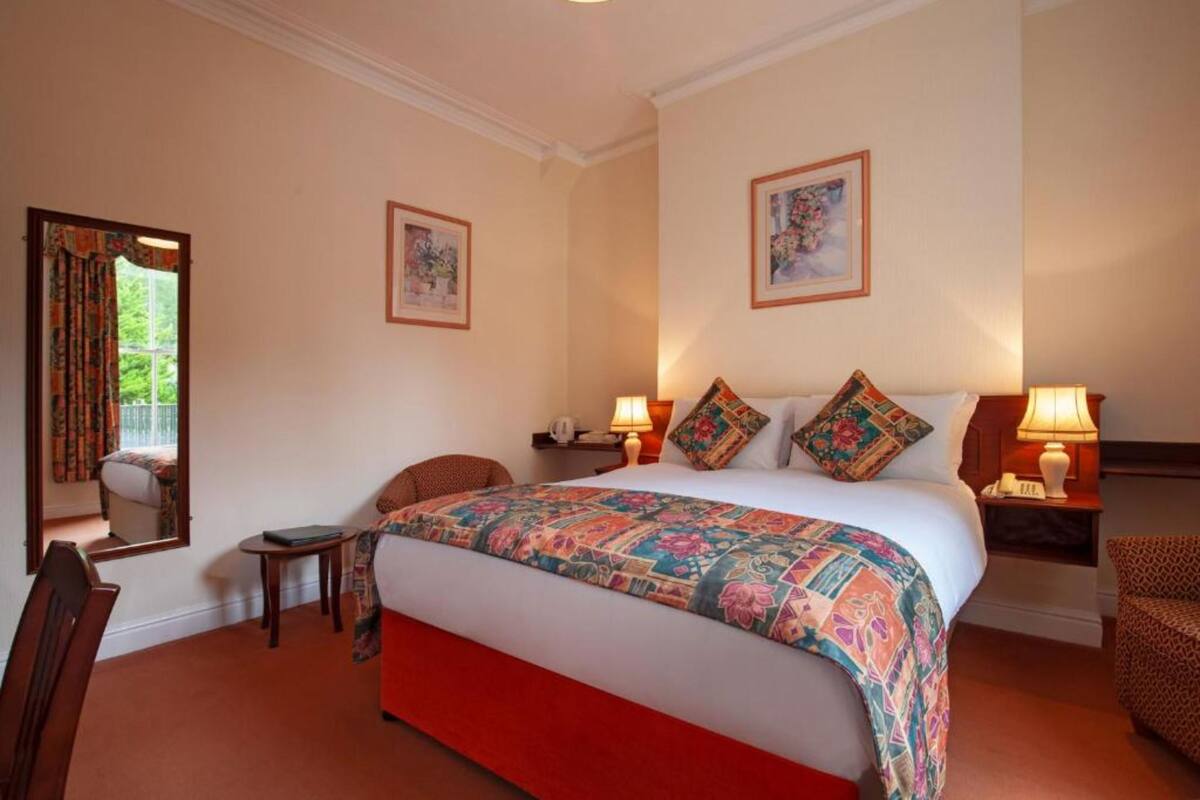 Quarto de hotel com uma cama grande, duas mesas de cabeceira com luminárias, uma cadeira, uma mesinha e uma janela com cortinas.