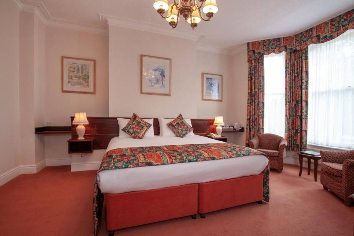 Quarto de hotel com uma cama grande, duas mesas de cabeceira com luminárias, uma cadeira, uma mesinha e uma janela com cortinas.