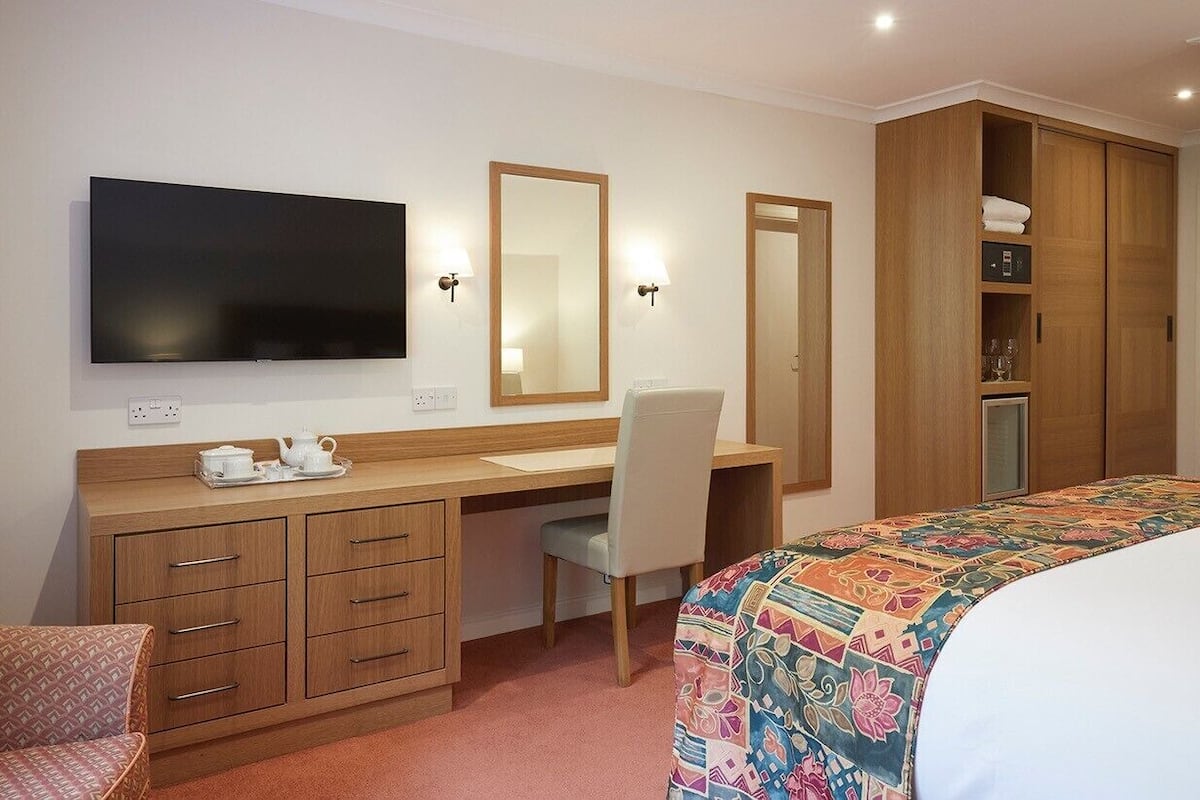 Quarto de hotel com uma cama, uma escrivaninha com cadeira, uma televisão, um espelho, um guarda-roupa e um pequeno armário com chá.